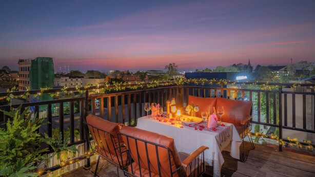 Terraço com mesa posta para dois, rodeado de cadeiras, com vista para a paisagem urbana ao entardecer.