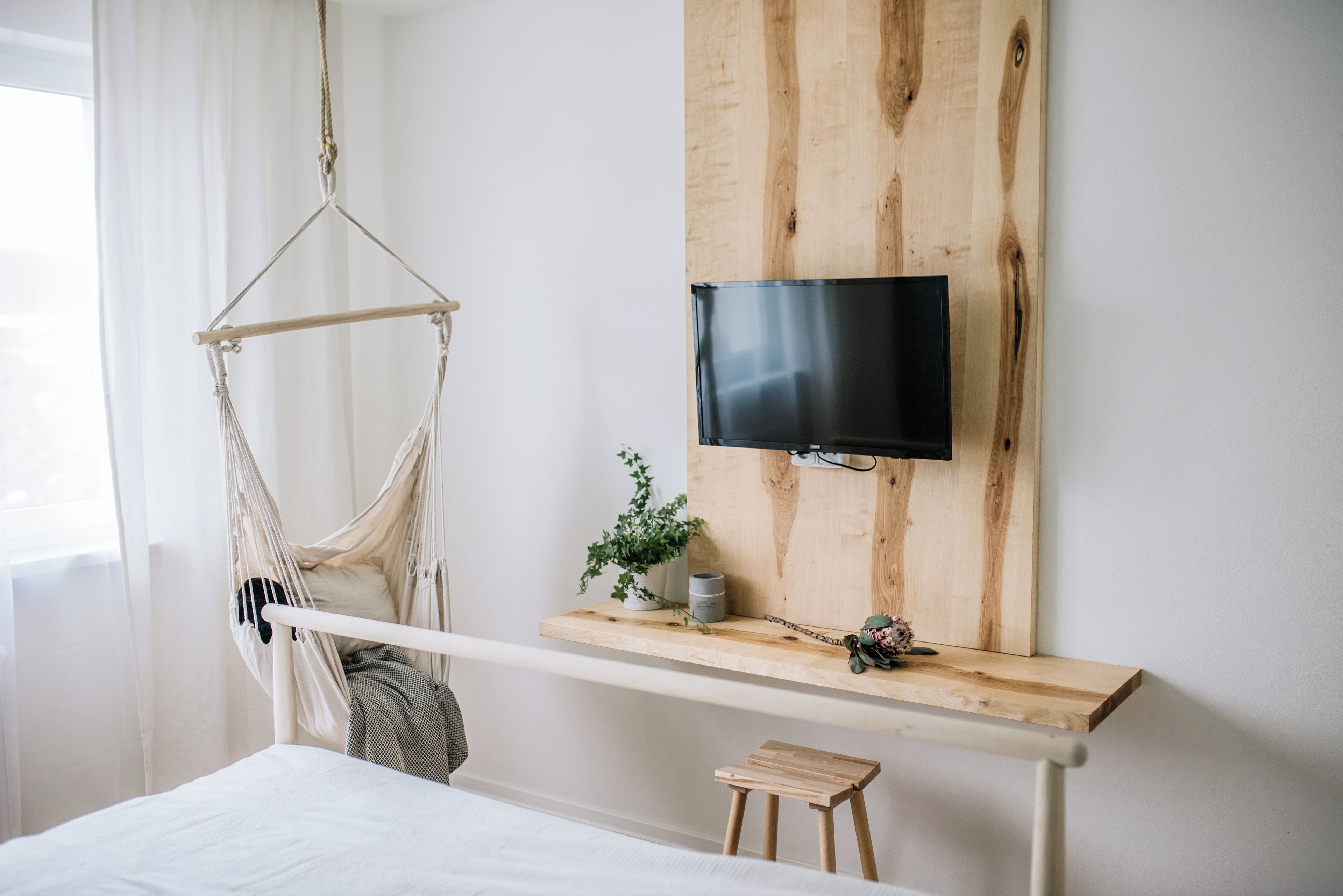 Quarto com cama, cadeira suspensa, televisão presa a uma tábua de madeira e um banquinho pequeno.
