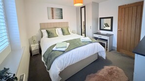 Um quarto bem arrumado, com cama, mesas de cabeceira, escrivaninha, espelho e porta.