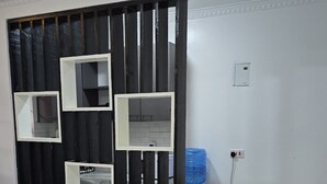 Quarto com divisória preta e branca com vários recortes quadrados, dispensador de água, frigorífico, cilindro de gás e fogão.