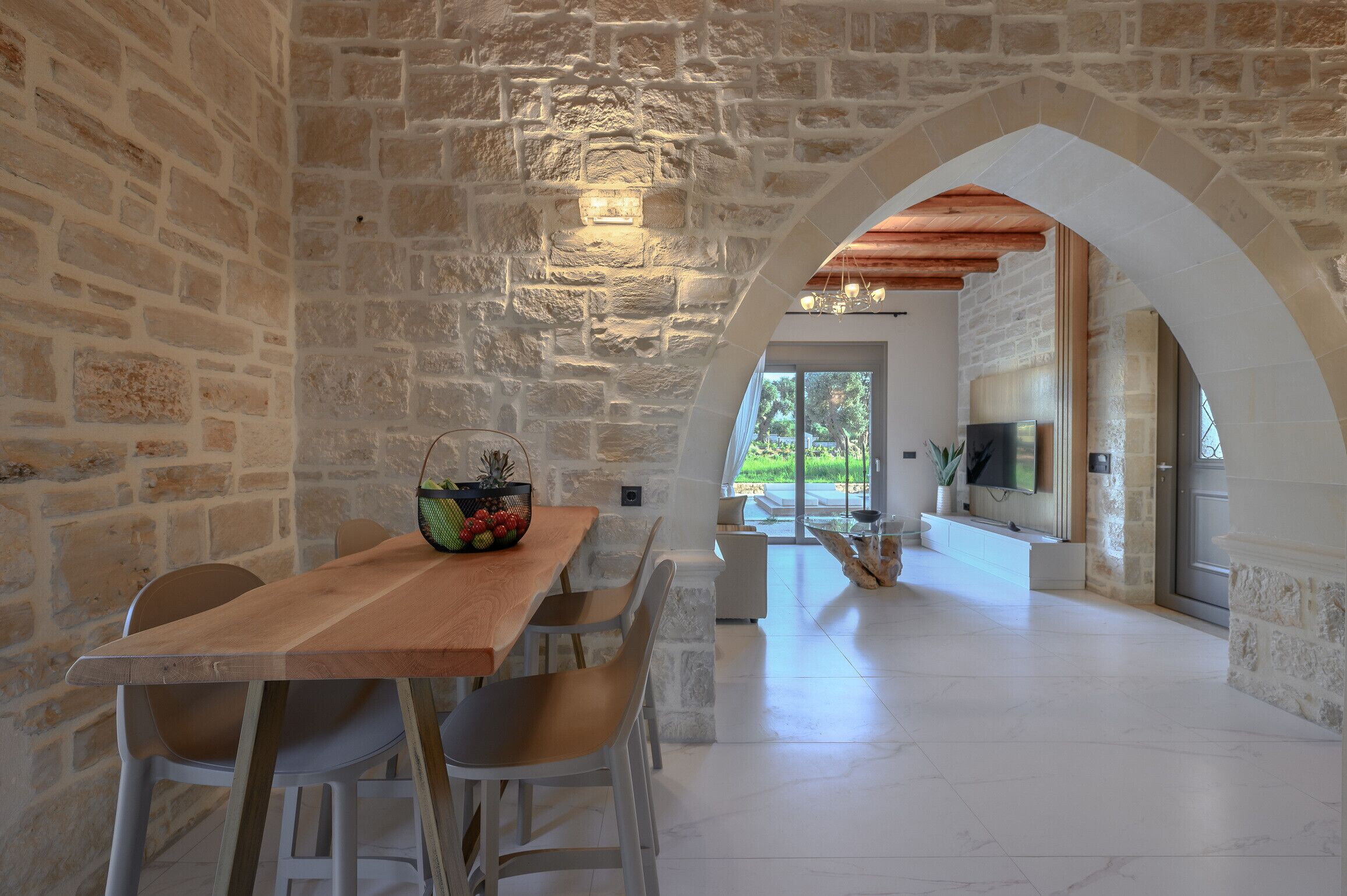 Um quarto com paredes de pedra, uma mesa de jantar de madeira e cadeiras, uma televisão montada na parede e vista para o exterior através de uma grande janela.