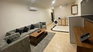 Sala de estar com sofá cinzento, mesa de centro de madeira e uma televisão pequena. É possível ver uma máquina de lavar e uma cozinha ao fundo.