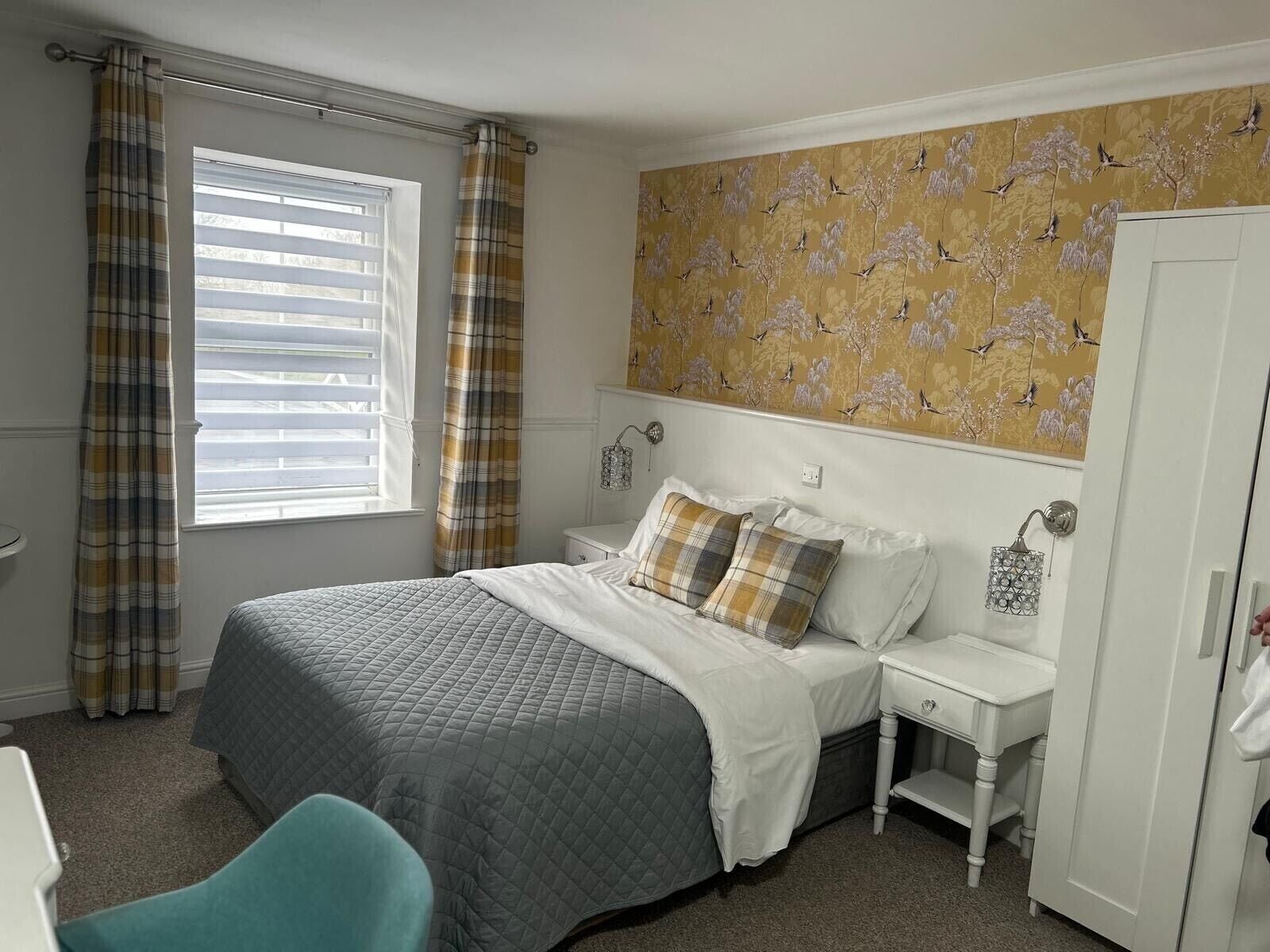 Quarto com cama, mesa de cabeceira, cadeira, guarda-roupa e janela com cortinas.