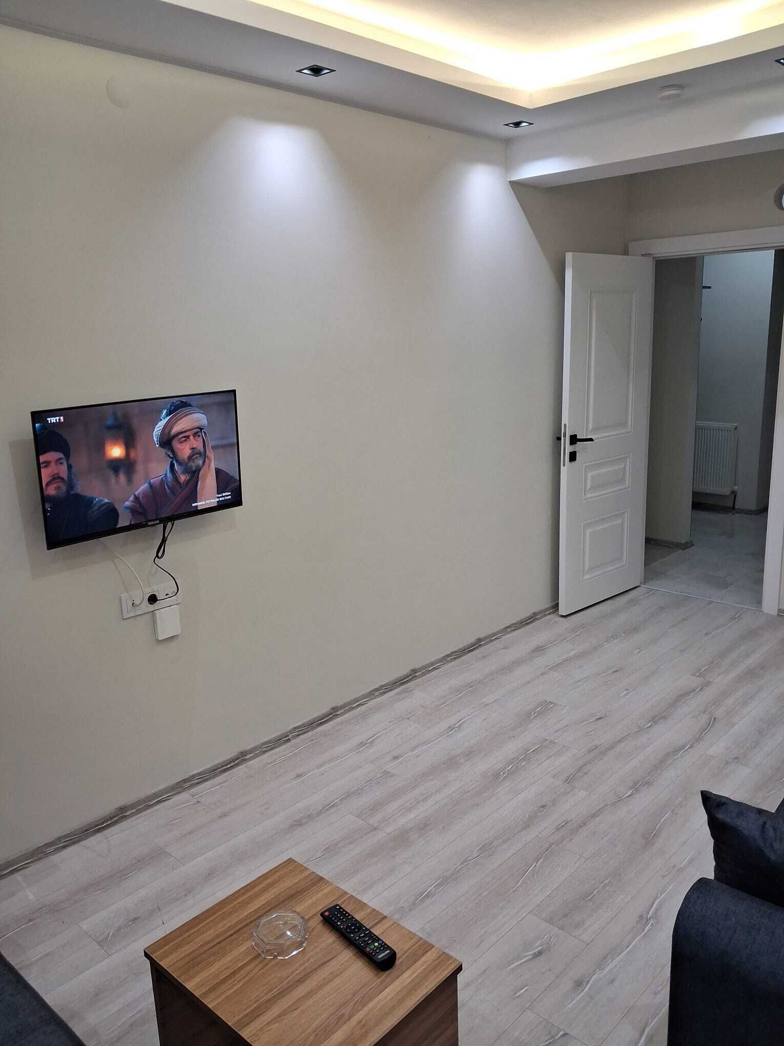 Uma sala de estar moderna com uma televisão de tela plana na parede, uma mesa de centro de madeira com controle remoto e um sofá de cor escura.