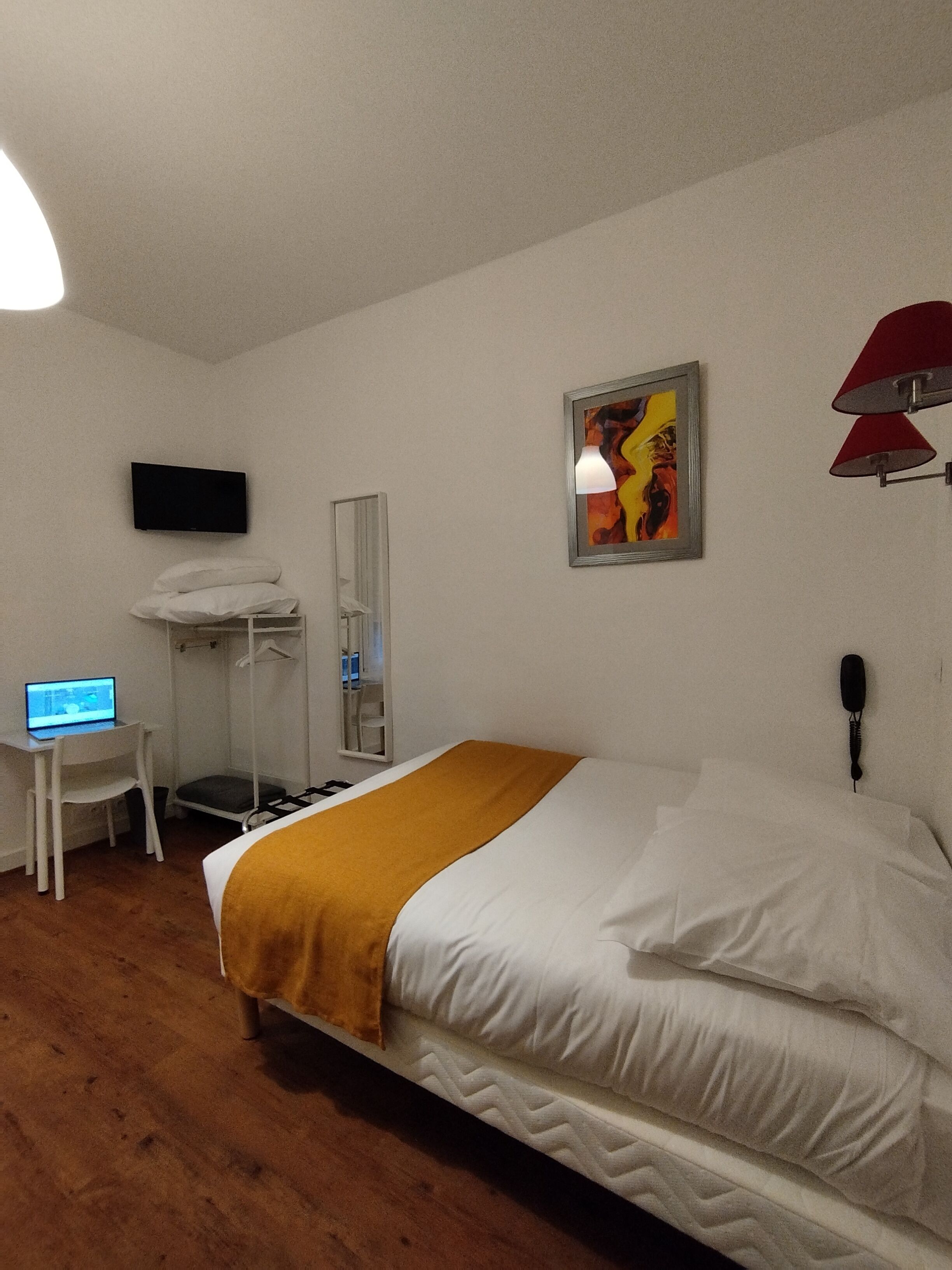 Quarto de hotel com cama, escrivaninha com laptop, televisão, quadro, espelho e telefone.