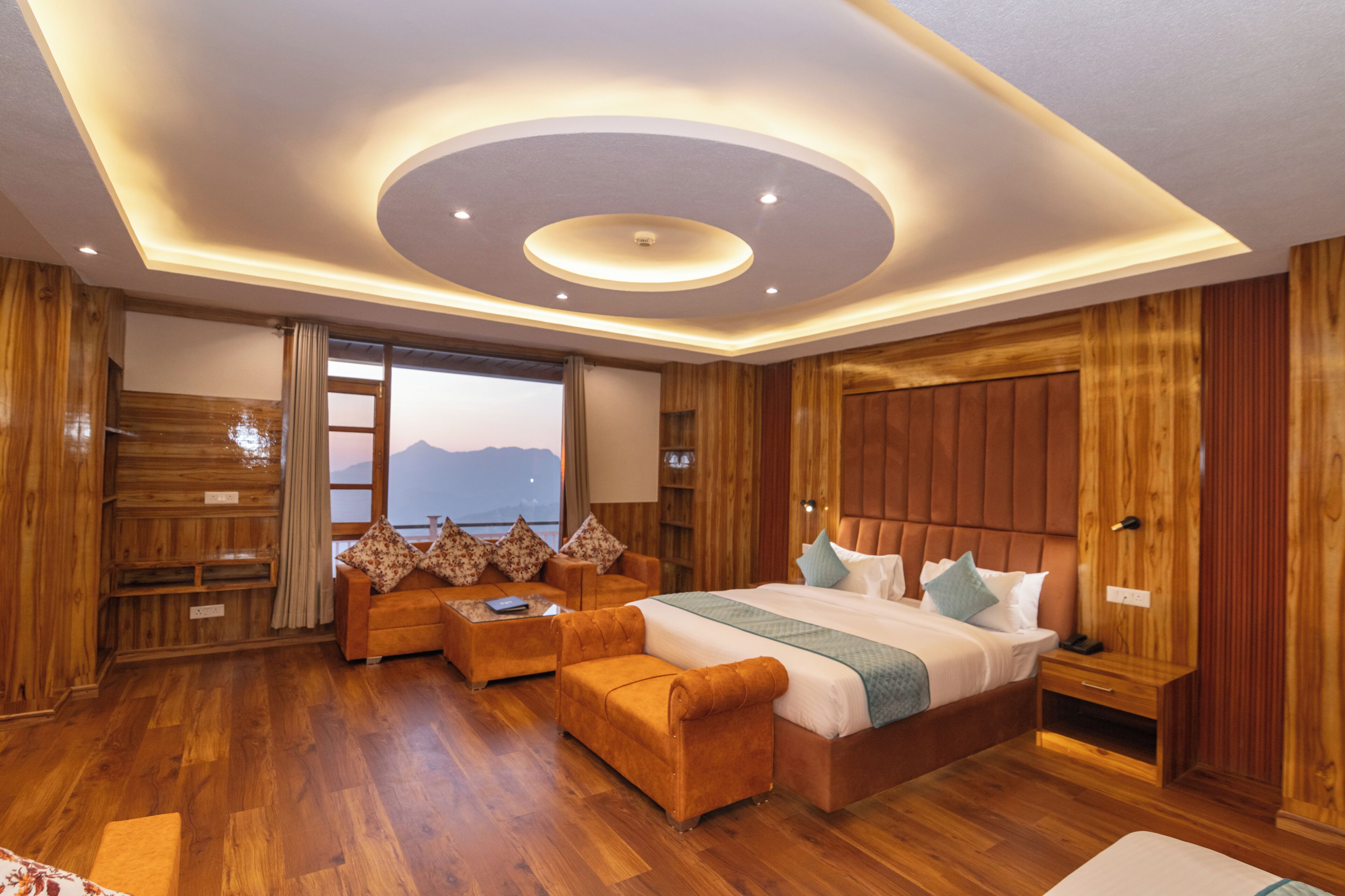 Quarto de hotel moderno com piso de madeira, uma cama grande com cabeceira, um sofá, uma mesa de cabeceira e vista para as montanhas pela janela.