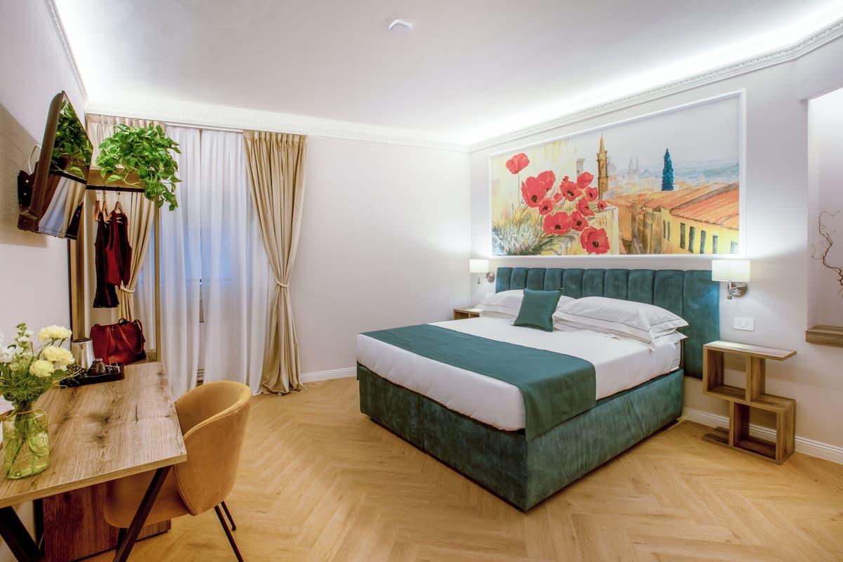 Quarto de hotel com cama, secretária com cadeira, planta e quadro na parede.