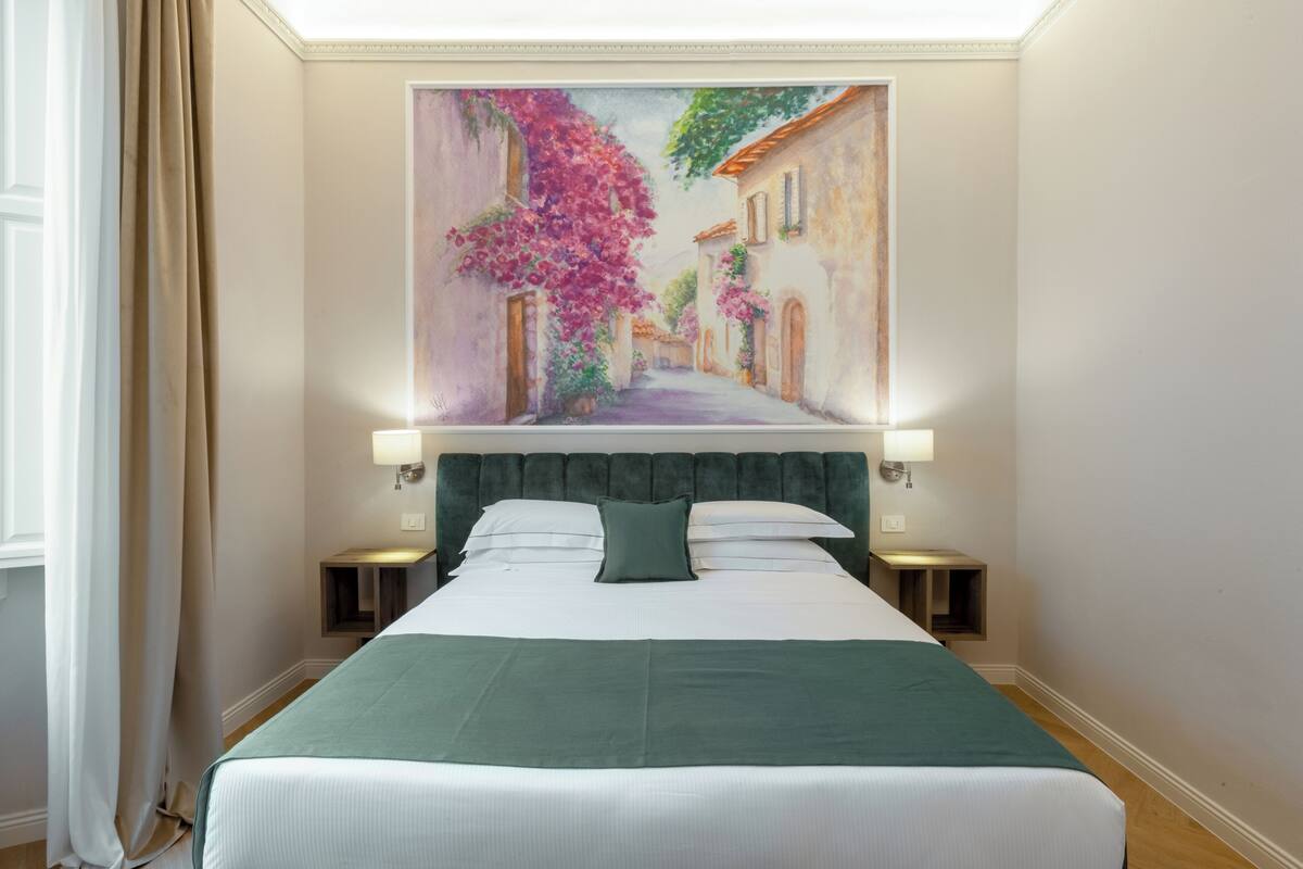 Quarto de hotel com uma cama grande, duas mesas de cabeceira, uma cabeceira e um quadro com uma cena de rua.