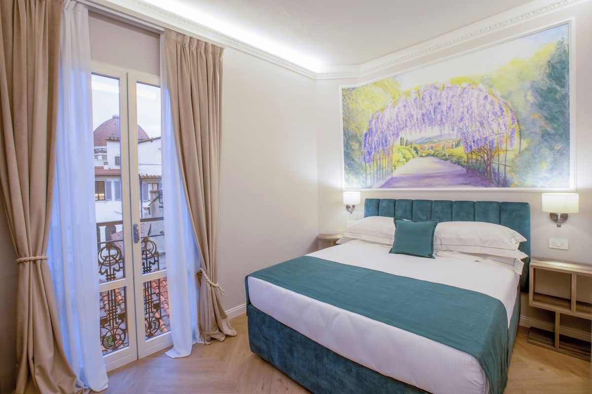 Quarto de hotel com uma cama grande, vista através de uma porta de vidro e um quadro na parede.