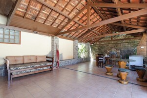 Um amplo alpendre coberto com teto de madeira, sof á, mesa de jantar, cadeiras e geladeira.