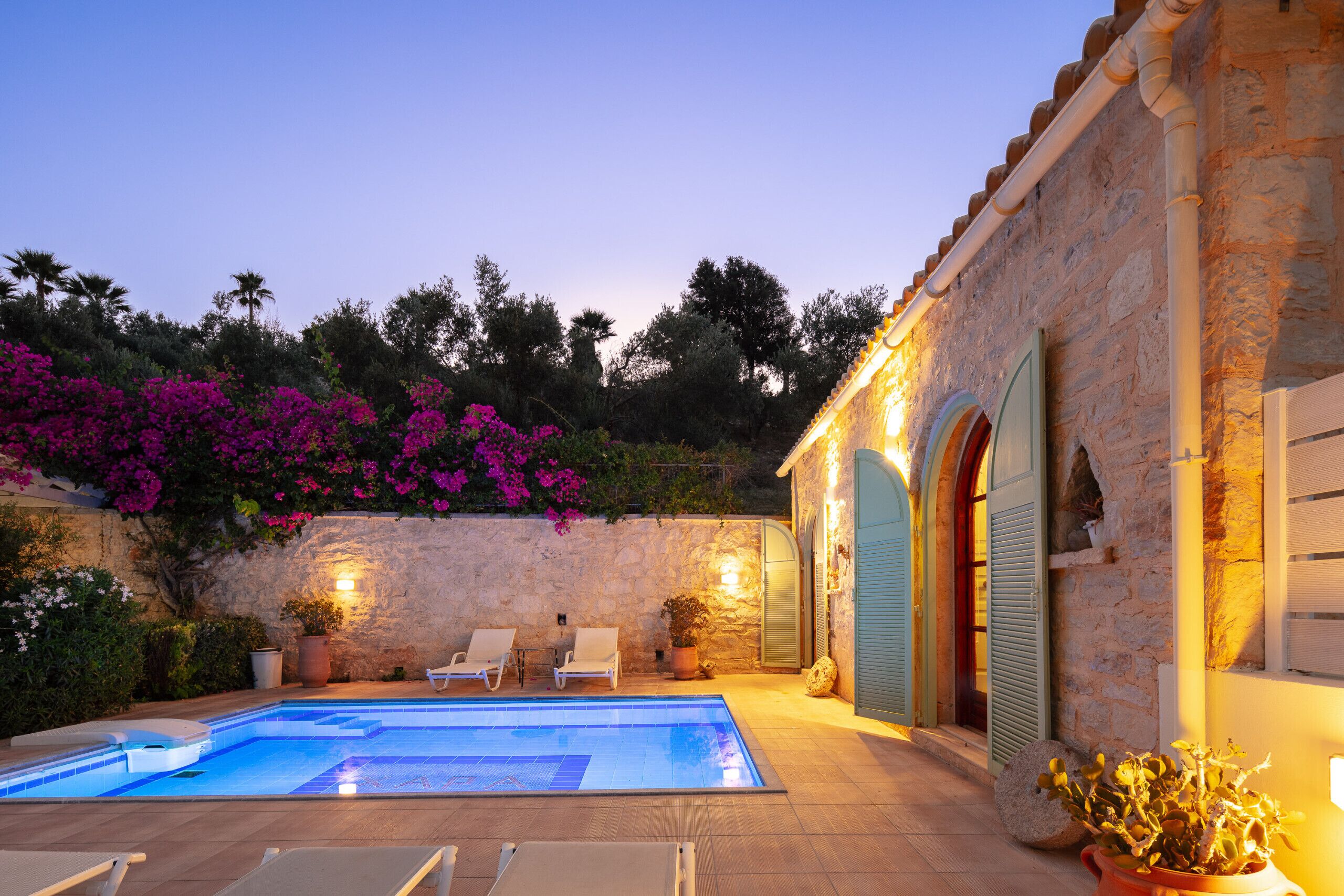 Piscina com espreguiçadeiras, parede de pedra e plantas em vasos.