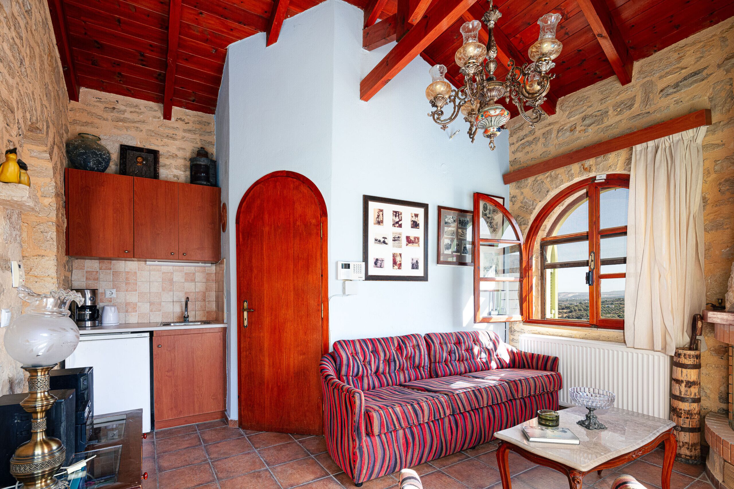 Uma sala de estar aconchegante com sofá, mesa de jantar, lustre e área de cozinha com armários de madeira e painel de azulejos.
