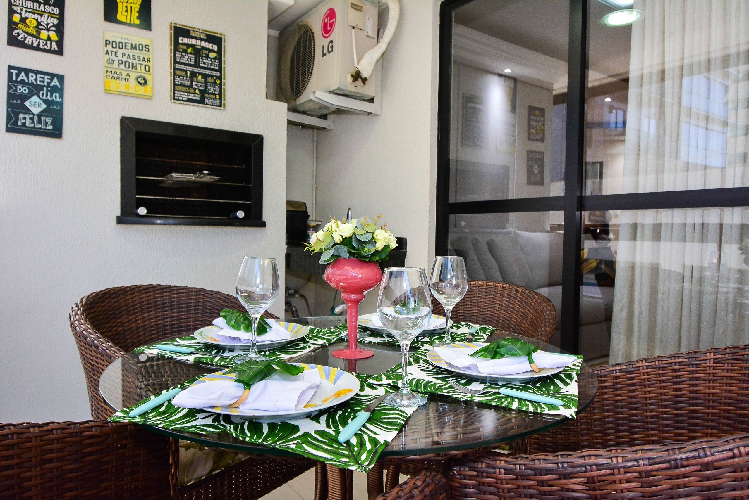 Mesa de jantar posta com guardanapos brancos, pratos e copos, rodeada por cadeiras de vime.