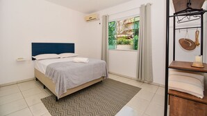 Quarto com cama, janela com cortinas, ar condicionado na parede e prateleira com toalhas.