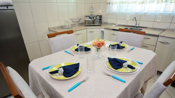 Mesa de jantar posta para quatro pessoas, com pratos amarelos, guardanapos azuis e copos de vidro.