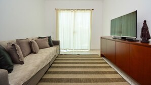 Uma sala de estar com sofá, televisão sobre um aparador de madeira e uma janela grande com cortinas leves.