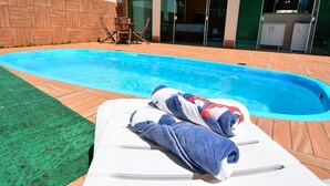 Piscina com espreguiçadeiras e toalhas, um deck de madeira e uma porta de vidro que dá para uma cozinha.