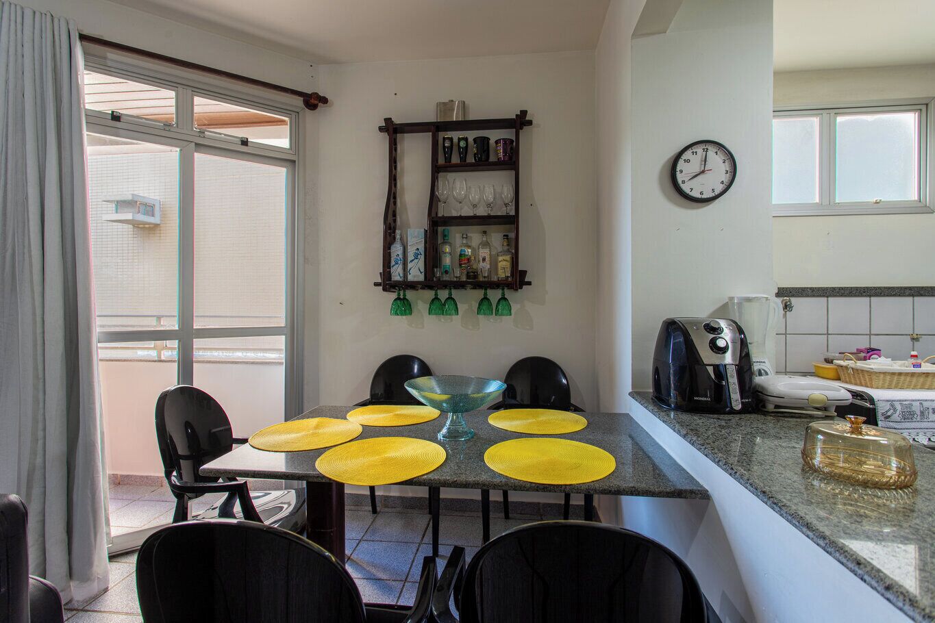 Uma área de refeições com uma mesa posta para quatro pessoas, cadeiras, um centro de mesa em uma tigela de vidro e uma cozinha com uma cafeteira e um relógio na parede.
