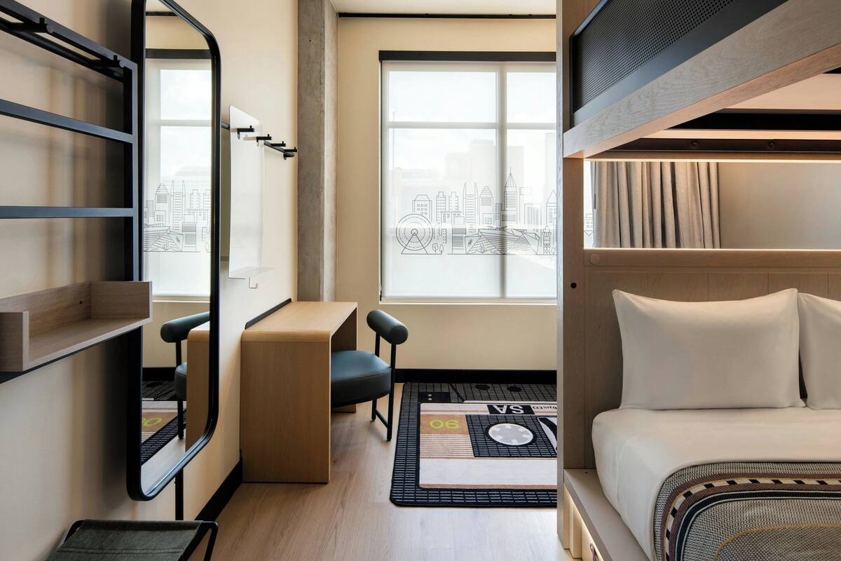 Quarto de hotel moderno com beliche, escrivaninha, cadeira, janela ampla com vista para a cidade e tapete estampado.