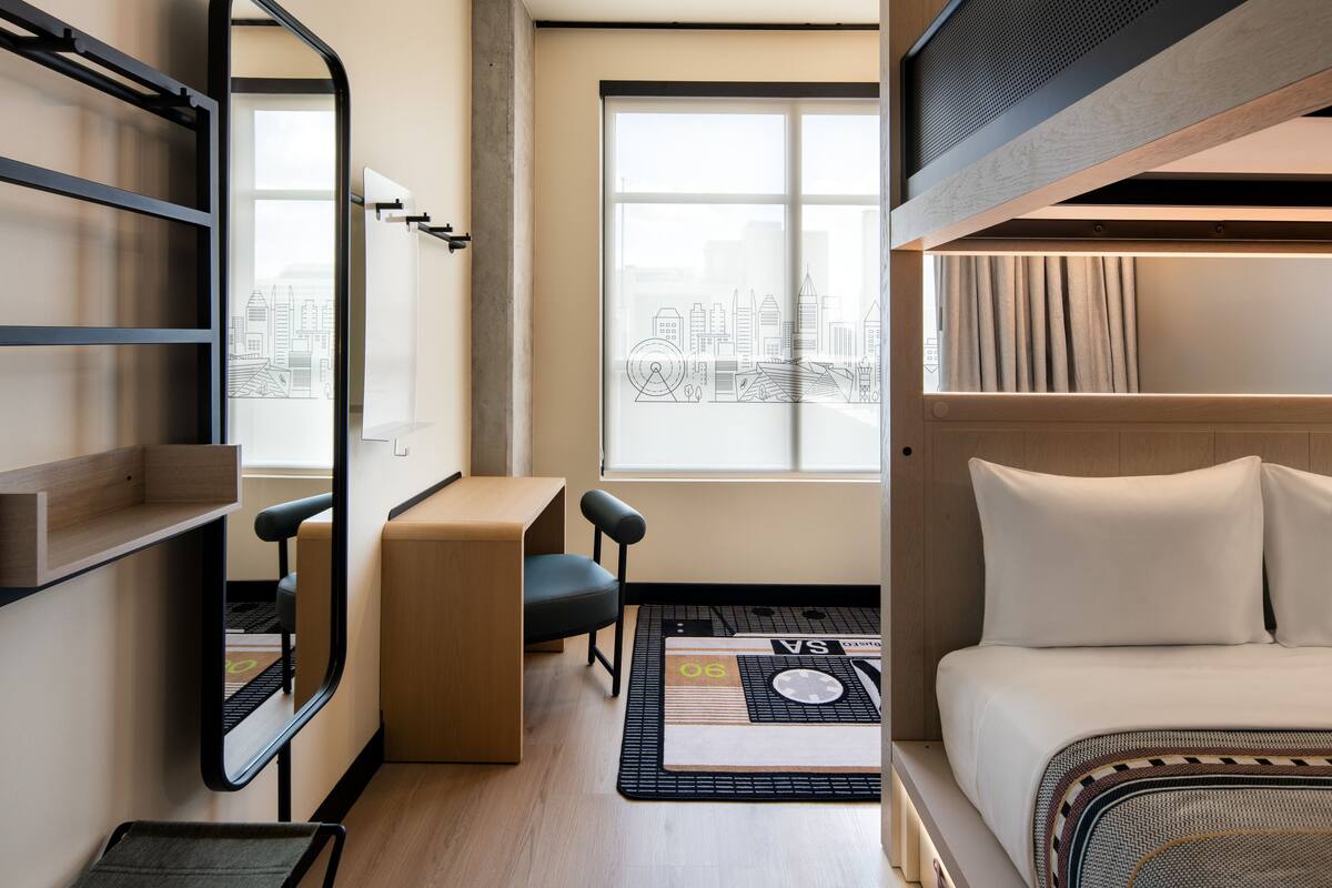 Quarto de hotel compacto com uma cama beliche, uma escrivaninha, uma cadeira, um espelho e vista para a cidade pela janela.