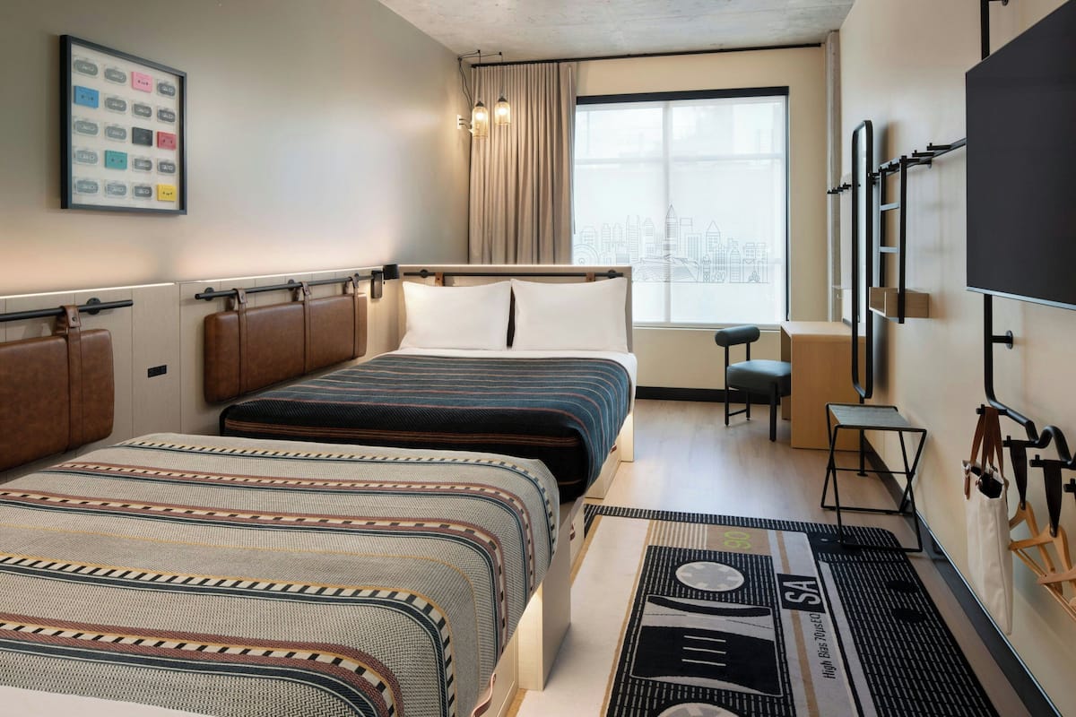 Quarto de hotel com duas camas, uma escrivaninha, uma cadeira, uma televisão e uma janela com vista.