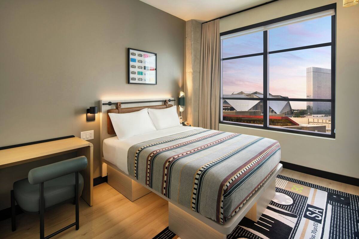 Quarto de hotel com cama, escrivaninha, cadeira, janela grande com vista para a cidade e quadro na parede.