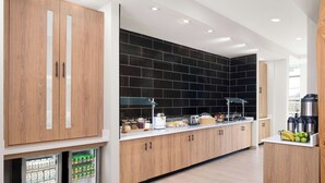 Cozinha moderna com armários de madeira, painel de azulejos pretos e bancada extensa com diversos eletrodomésticos e alimentos.