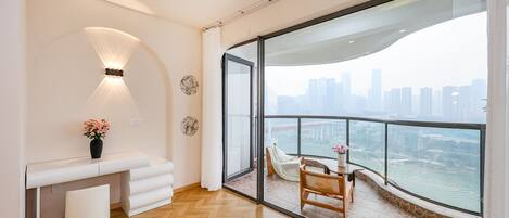 Quarto moderno com uma janela grande, uma escrivaninha branca, uma cadeira e vista para a cidade e rio.