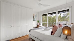 Quarto com cama, guarda-roupa branco, escrivaninha e janela com vista para árvores.