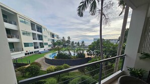 Uma varanda com vista para uma piscina, palmeiras e um edifício moderno.