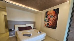 Quarto de hotel moderno com cama grande, cabeceira, mesa de cabeceira, quadro na parede e uma porta à direita.