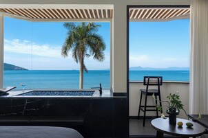 Uma varanda com vista para o mar e uma palmeira, uma pequena mesa com uma planta e uma cadeira.