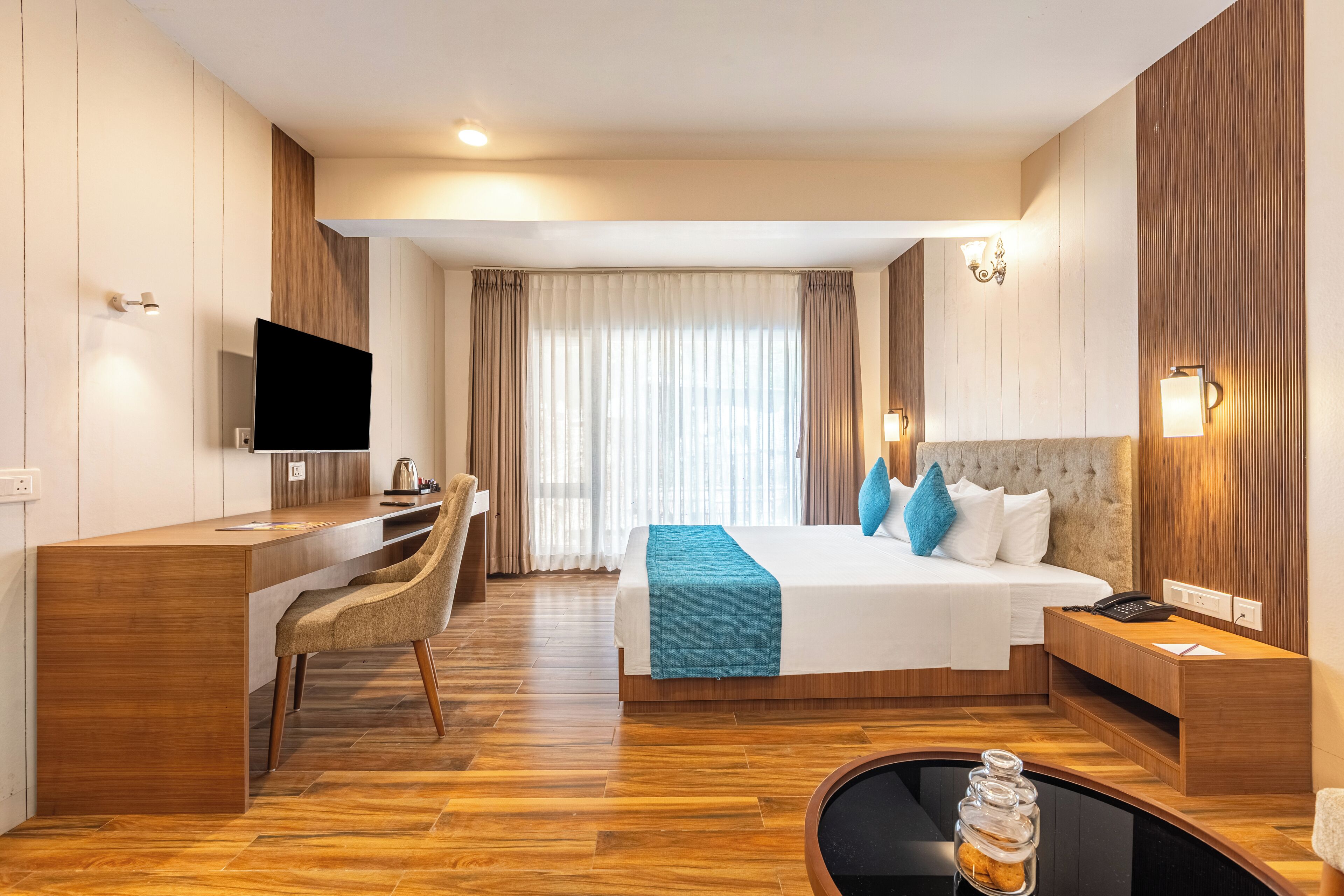 Quarto de hotel com uma escrivaninha de madeira, uma cama com manta azul, uma televisão na parede e uma máquina de café.