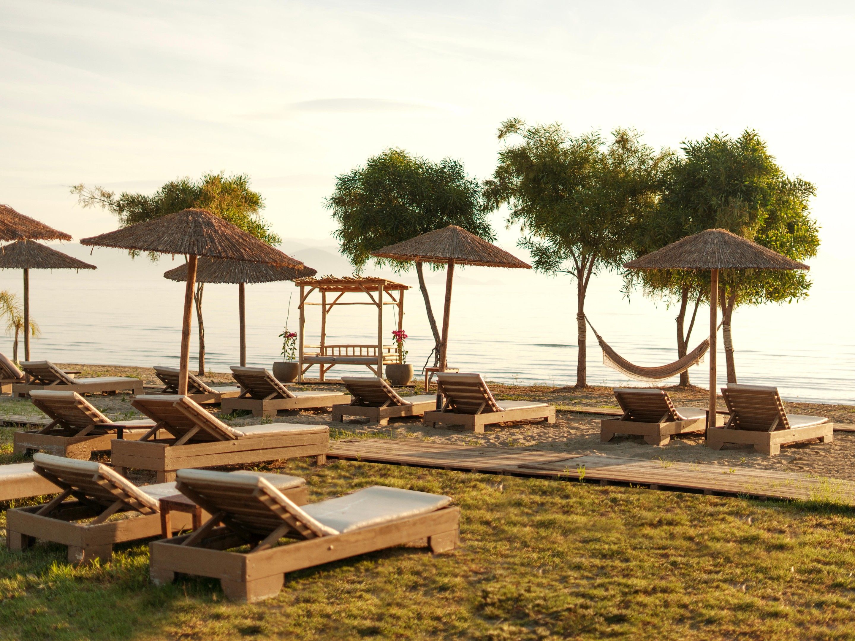Uma área de lounge à beira-mar com espreguiçadeiras de madeira e guarda-sóis de palha, um balanço e um deck de madeira.