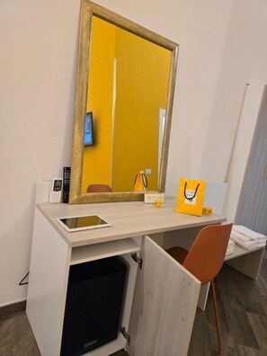 Quarto de hotel com escrivaninha, espelho, cadeira, um eletrodoméstico preto pequeno e uma bolsa amarela sobre a escrivaninha.