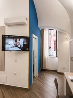 Quarto de hotel moderno com televisão de tela plana na parede, piso de madeira, parede azul como destaque e uma janela com cortinas.