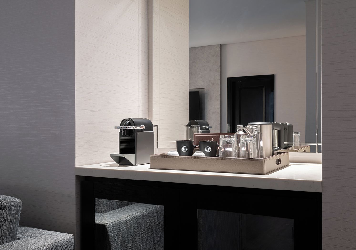 Quarto de hotel moderno com design minimalista, apresentando um espaço para café com cafeteira, xícaras e um tabuleiro, e um espelho grande refletindo o interior do quarto.