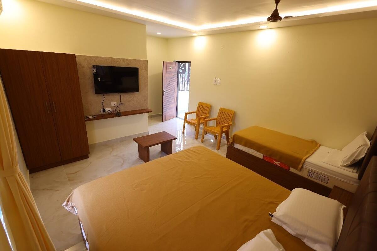 Quarto de hotel com cama, televisão presa na parede, escrivaninha, duas cadeiras e guarda-roupa.