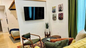 Uma sala de estar com um sofá amarelo, uma cadeira de madeira, um tapete estampado, uma televisão de tela plana na parede e um pequeno vaso com planta.