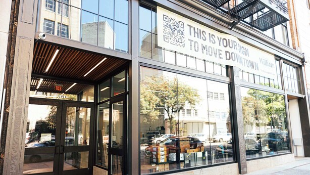 Fachada com grandes janelas e um letreiro com a frase 'ESTE É O SEU CONVITE PARA MUDAR PARA O CENTRO.'