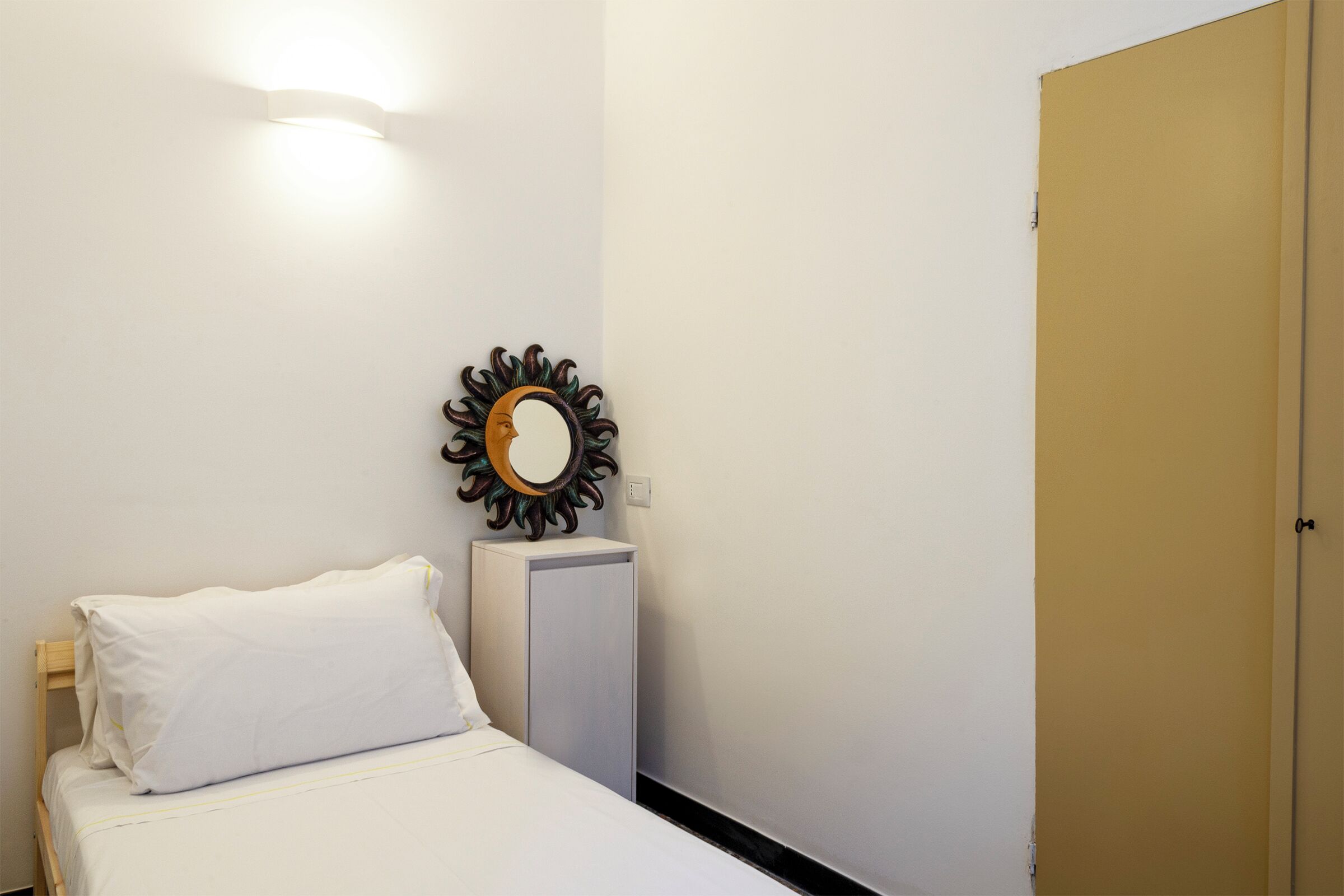 Quarto pequeno e bem organizado, com uma cama de solteiro, mesa de cabeceira, espelho e guarda-roupa.