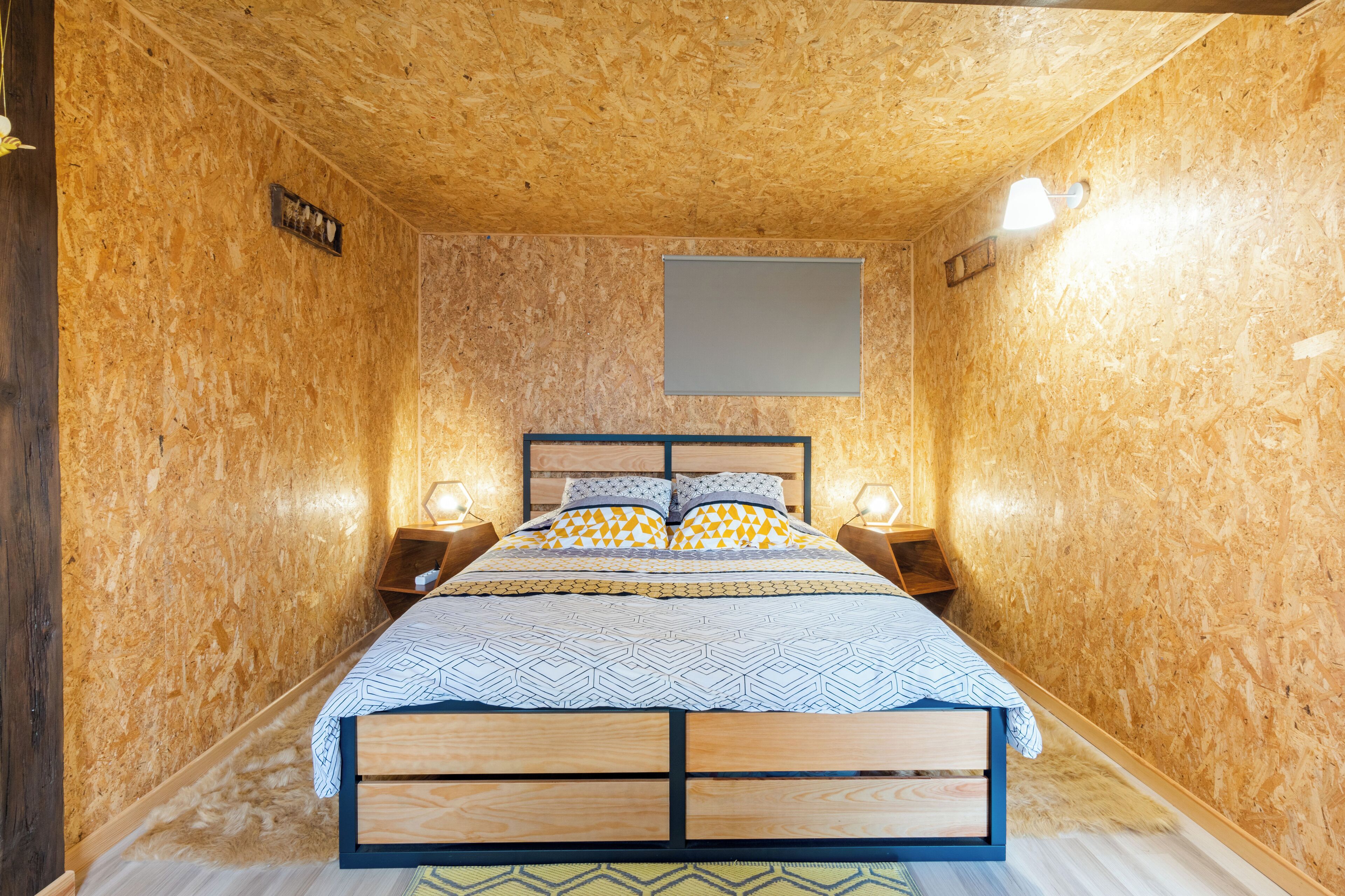 Quarto com cabeceira de madeira, cama com roupa de cama estampada, duas mesas de cabeceira com luminárias e luminária fixada na parede.