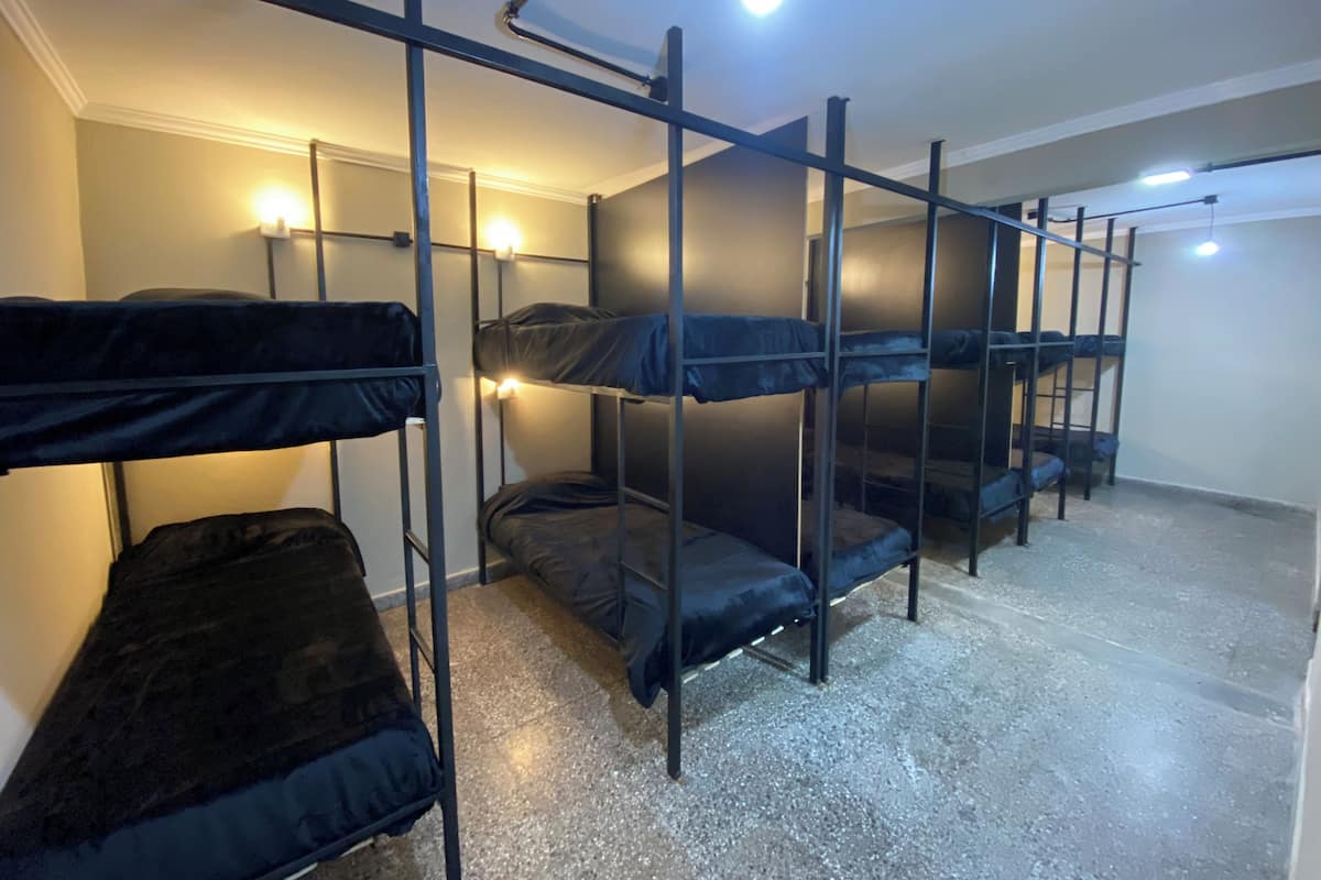 Quarto em dormitório com beliches dispostos em duas fileiras, uma luminária no teto e uma parede sem decoração.