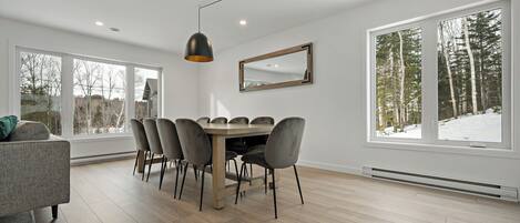 Uma área de refeições moderna com uma mesa e cadeiras de madeira, janelas amplas com vista para árvores, e um lustre.
