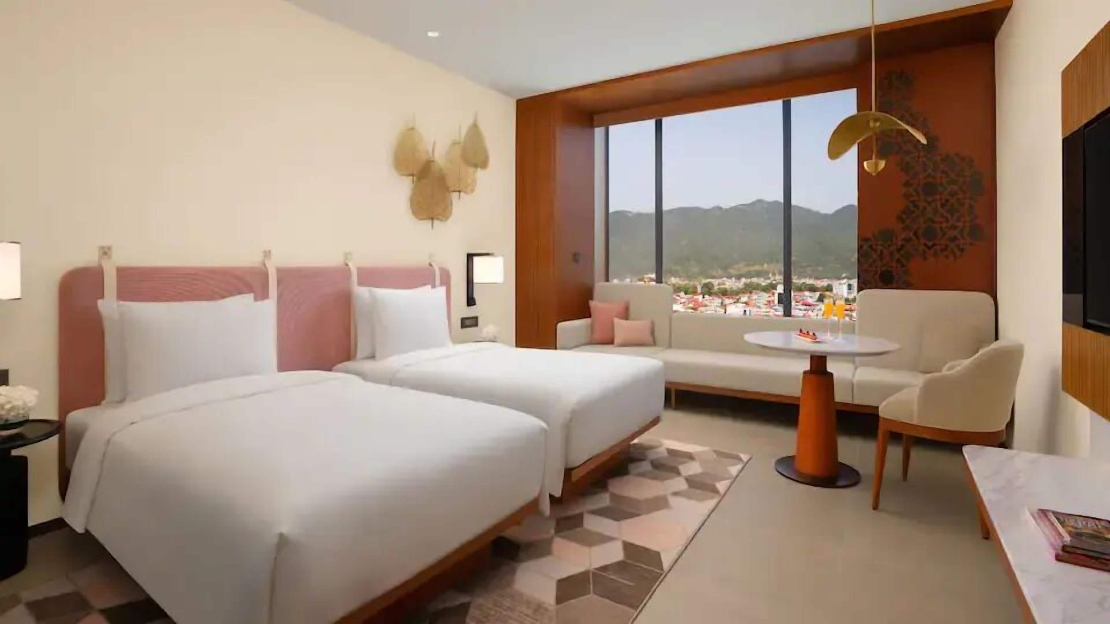 Quarto de hotel com duas camas, um sofá, uma mesa pequena e uma grande janela com vista para as montanhas.