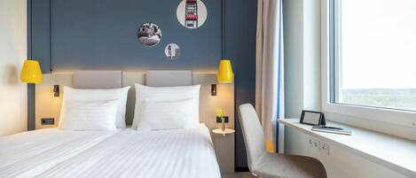 Quarto de hotel com uma cama, duas luminárias de cabeceira, uma escrivaninha com cadeira, uma janela e decoração na parede.