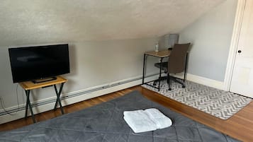 Um quarto pequeno e acolhedor, com uma cama, televisão sobre um suporte de madeira, uma escrivaninha com cadeira e um tapete estampado.
