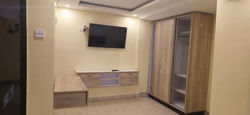 Quarto com televisão de tela plana na parede, guarda-roupa de madeira e pequena escrivaninha com gavetas.