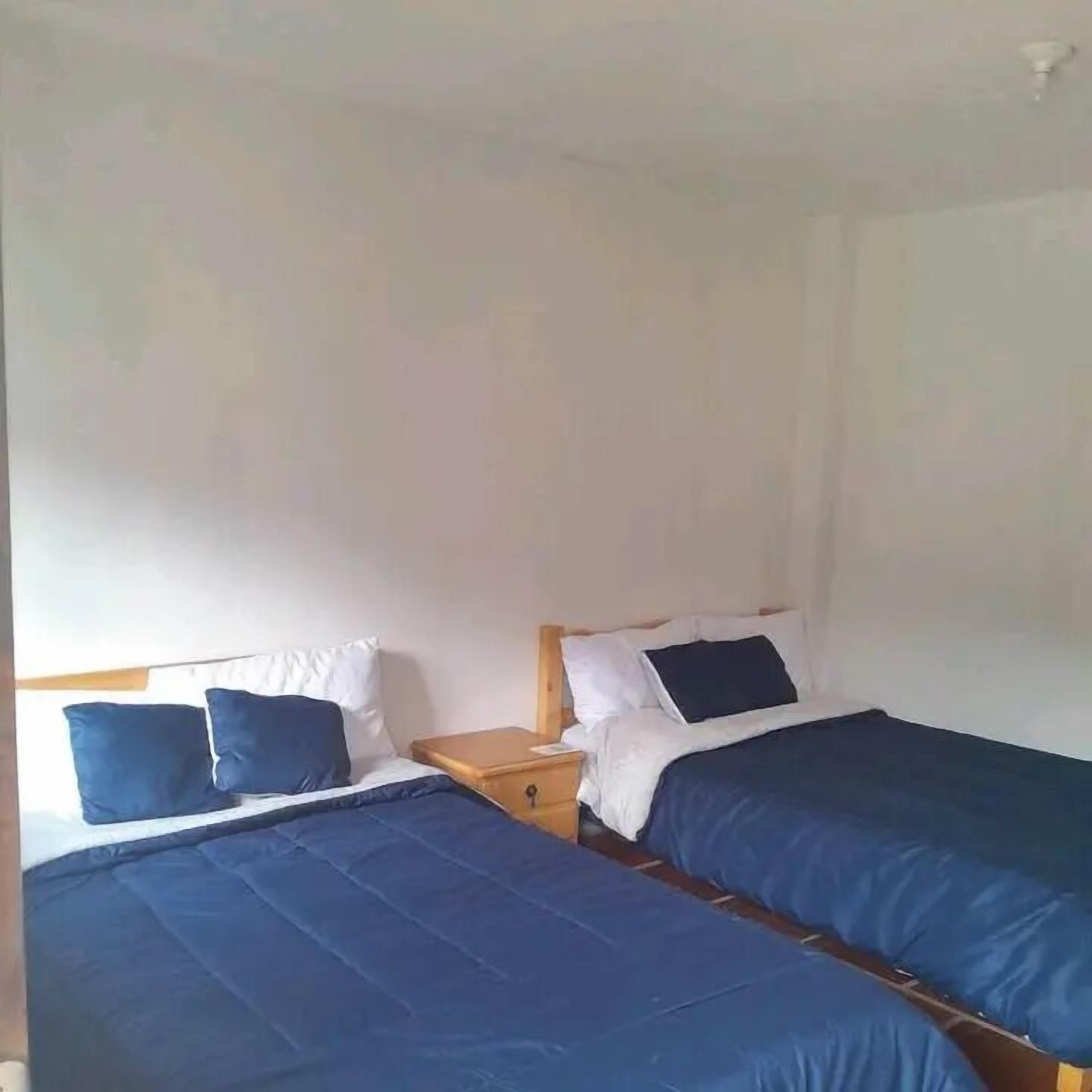 Quarto com duas camas individuais, cada uma com cabeceira de madeira e roupa de cama azul. Há uma mesinha de cabeceira de madeira entre as camas e um pequeno móvel com gavetas de madeira à direita.