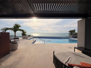 Uma área de piscina no terraço com vista para o mar, um espaço de descanso com assentos e uma palmeira.